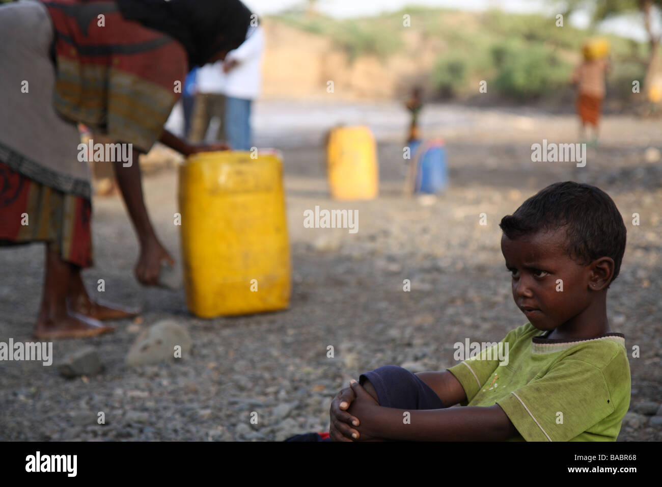 Child by a riverbank with women collecting water in the background ...