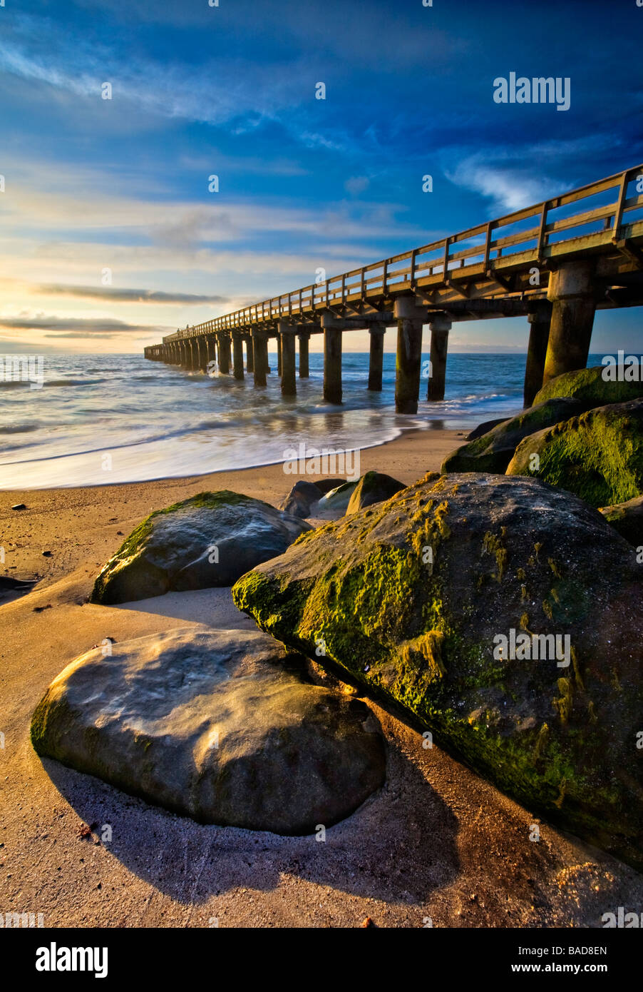 A large wooden pier at sunset with rocks covered in green moss all lit ...