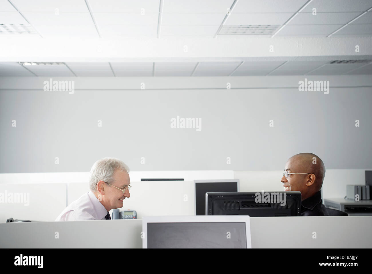 Two businessmen sitting face to face in office cubicle, smiling, side ...