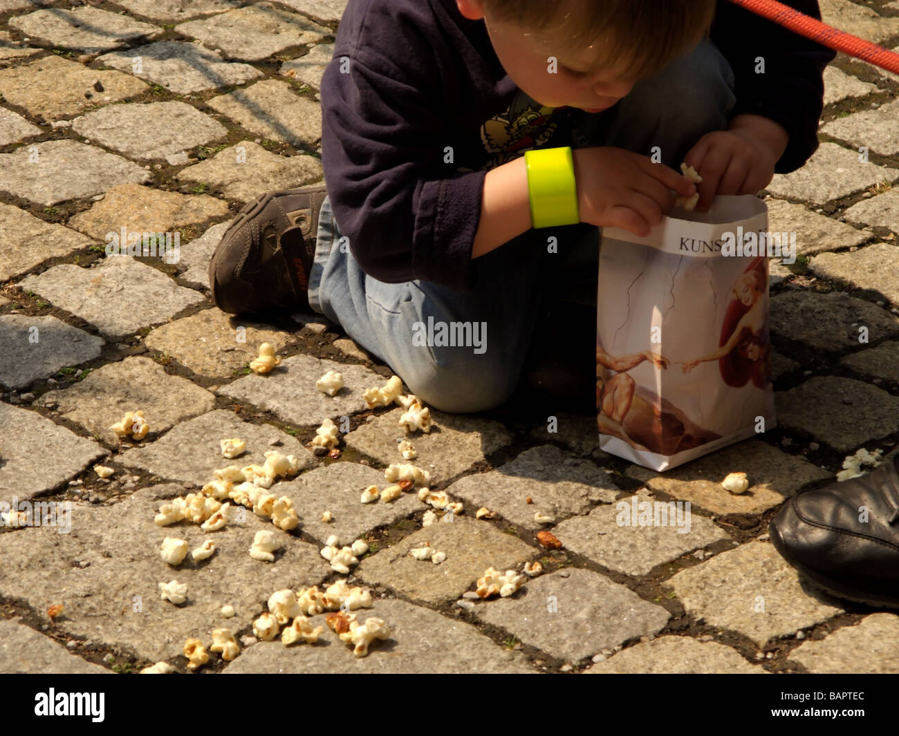Child Picking Up Popcorn from Cobblestone Pavement and Placing it into ...