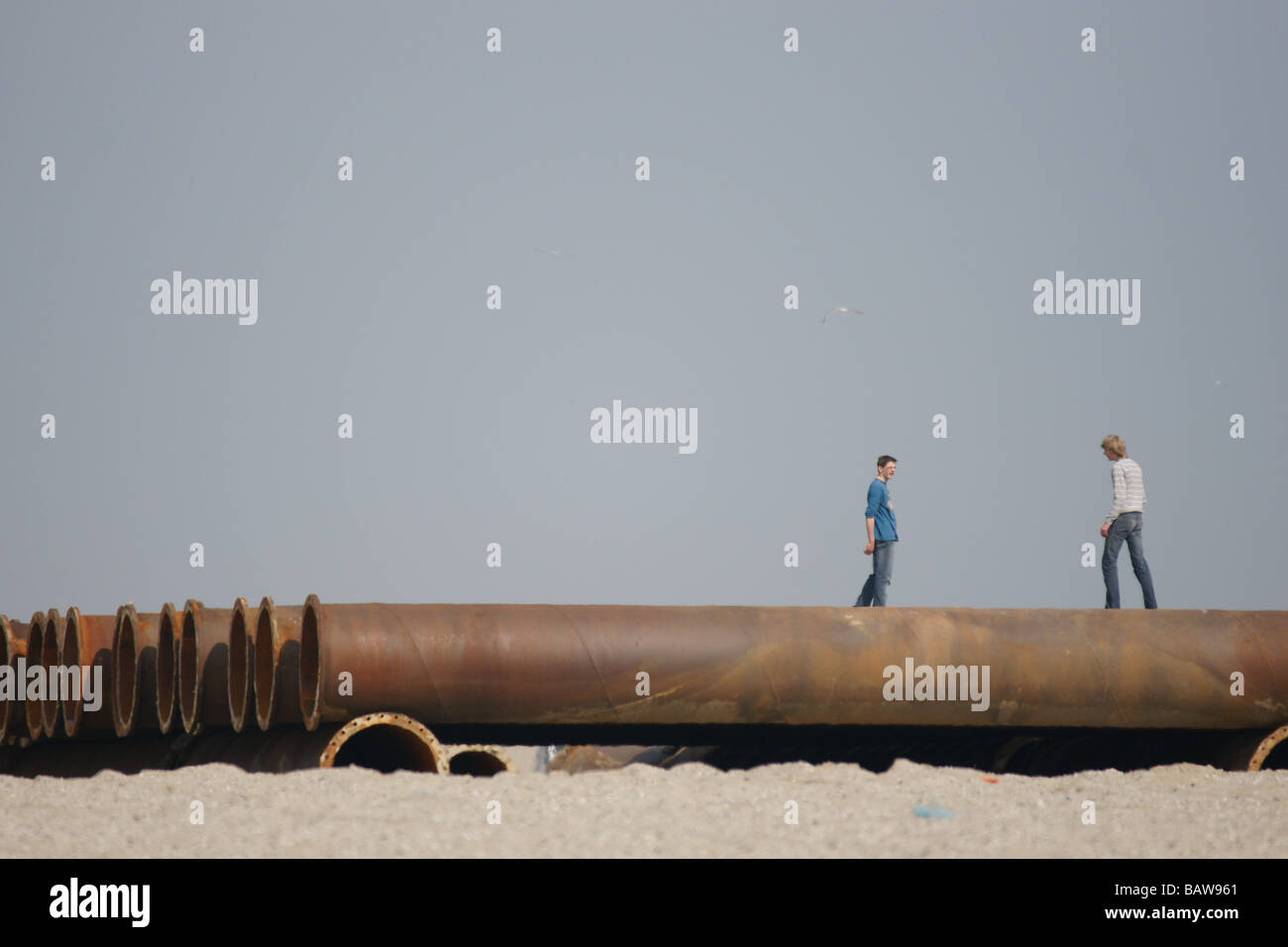 People walking on huge steel pipes pipeline in sand beach under blue ...