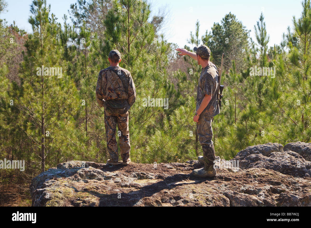 TWO HUNTERS IN CAMO STAND ON A ROCKY CLIFF LOOKING OVER THE TOP OF A ...