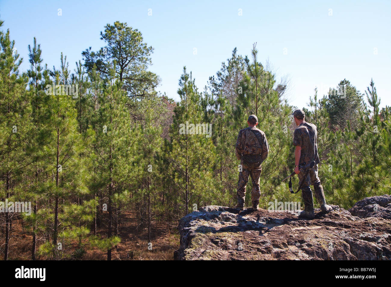TWO HUNTERS IN CAMO STAND ON A ROCKY CLIFF LOOKING OVER THE TOP OF A ...