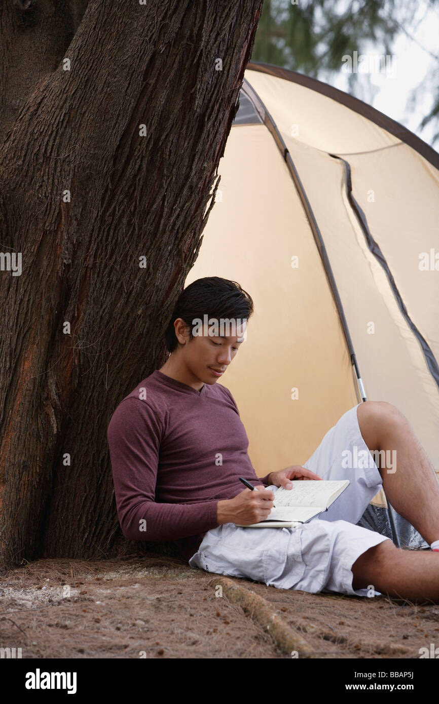 Man leaning against tree writing in journal, notebook, with tent in ...