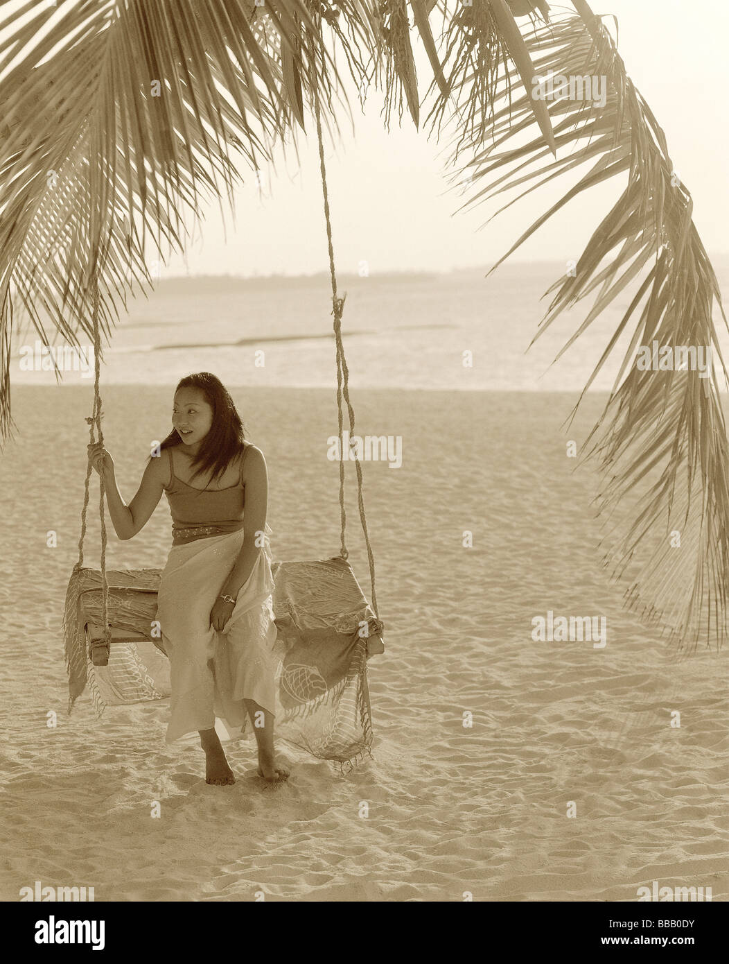 Woman sitting on swing under coconut tree looking off camera, sea in ...