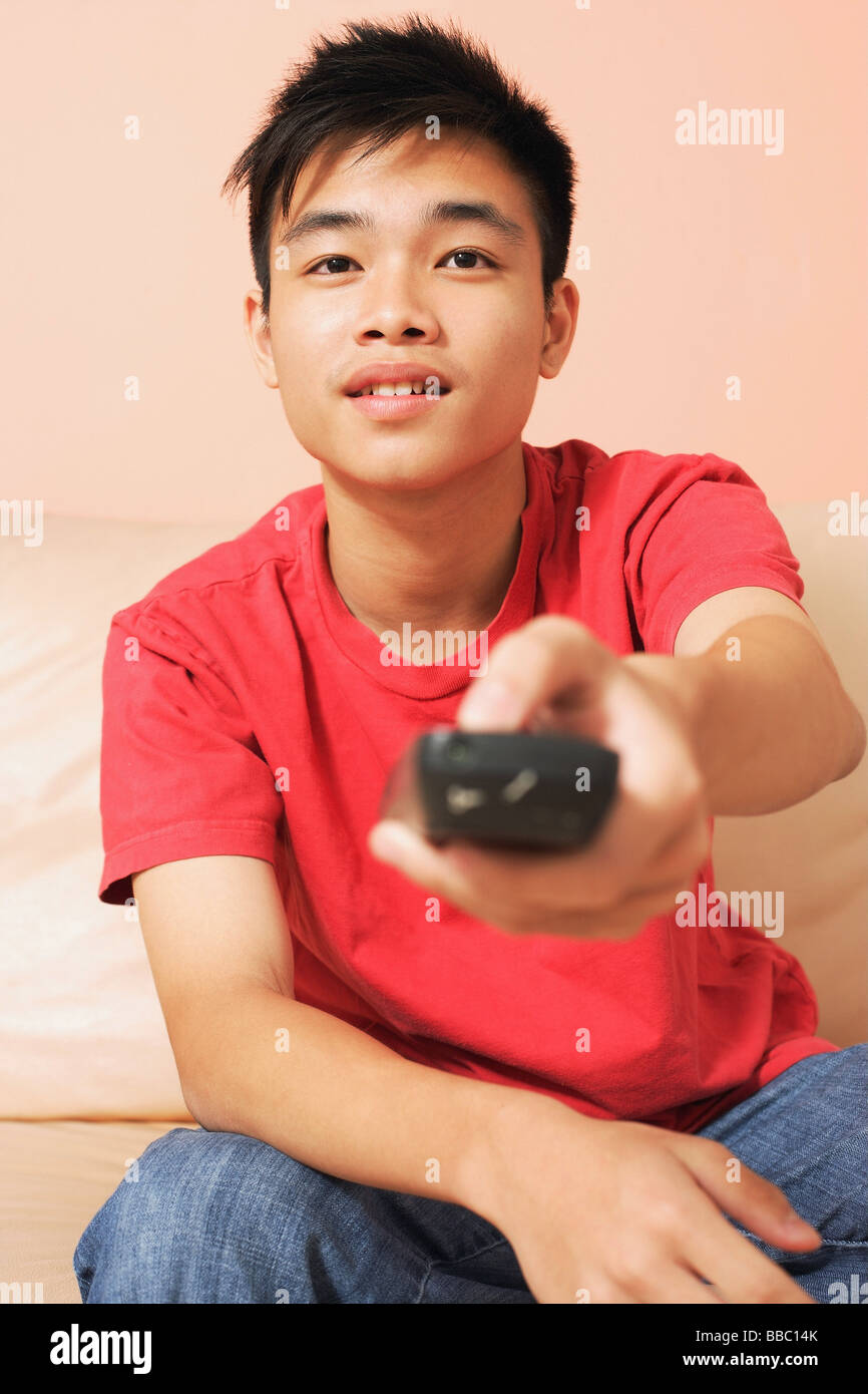 Teenage boy sitting on sofa, pointing remote control towards camera ...