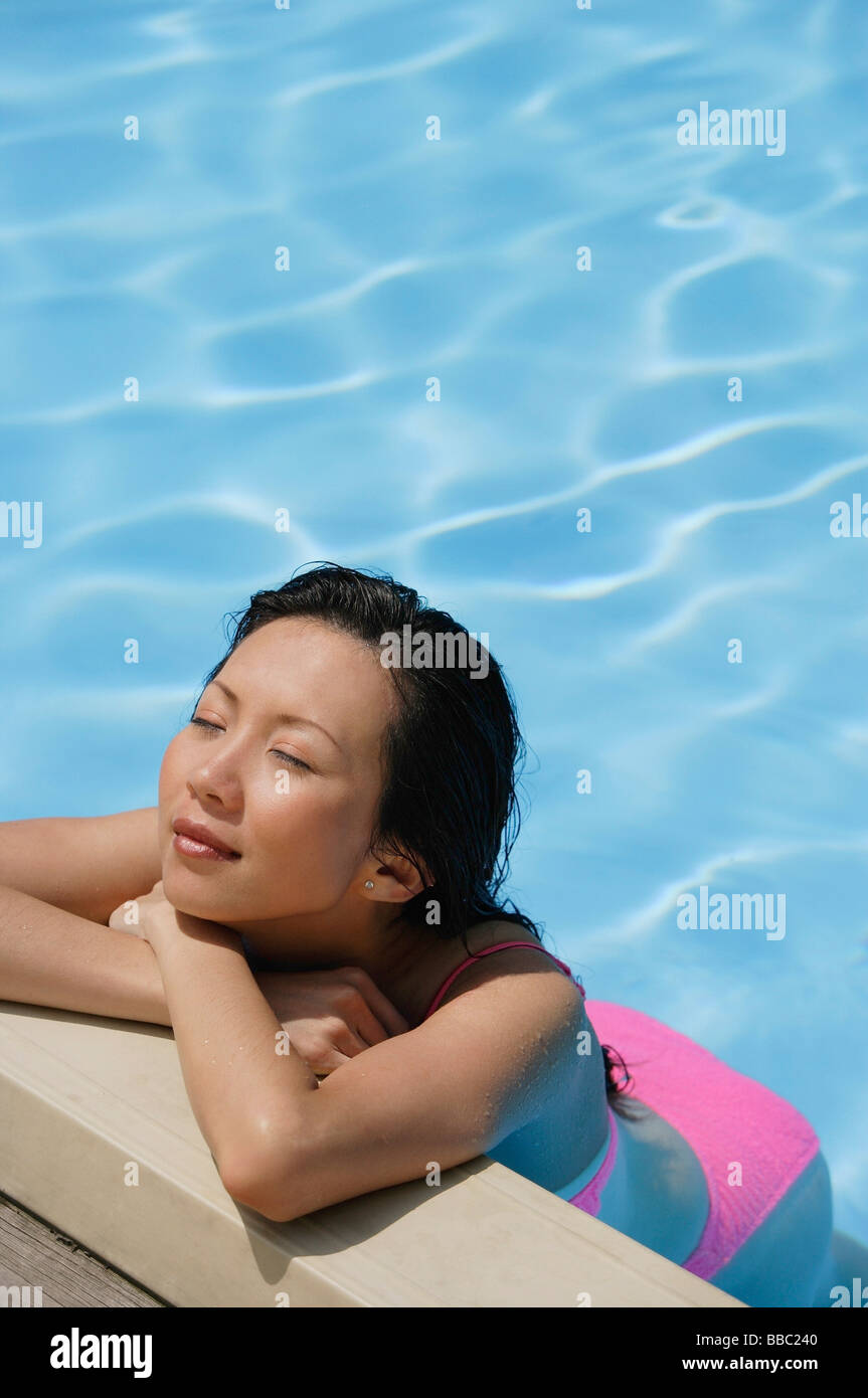 Woman leaning at the edge of swimming pool, arms crossed, eyes closed ...