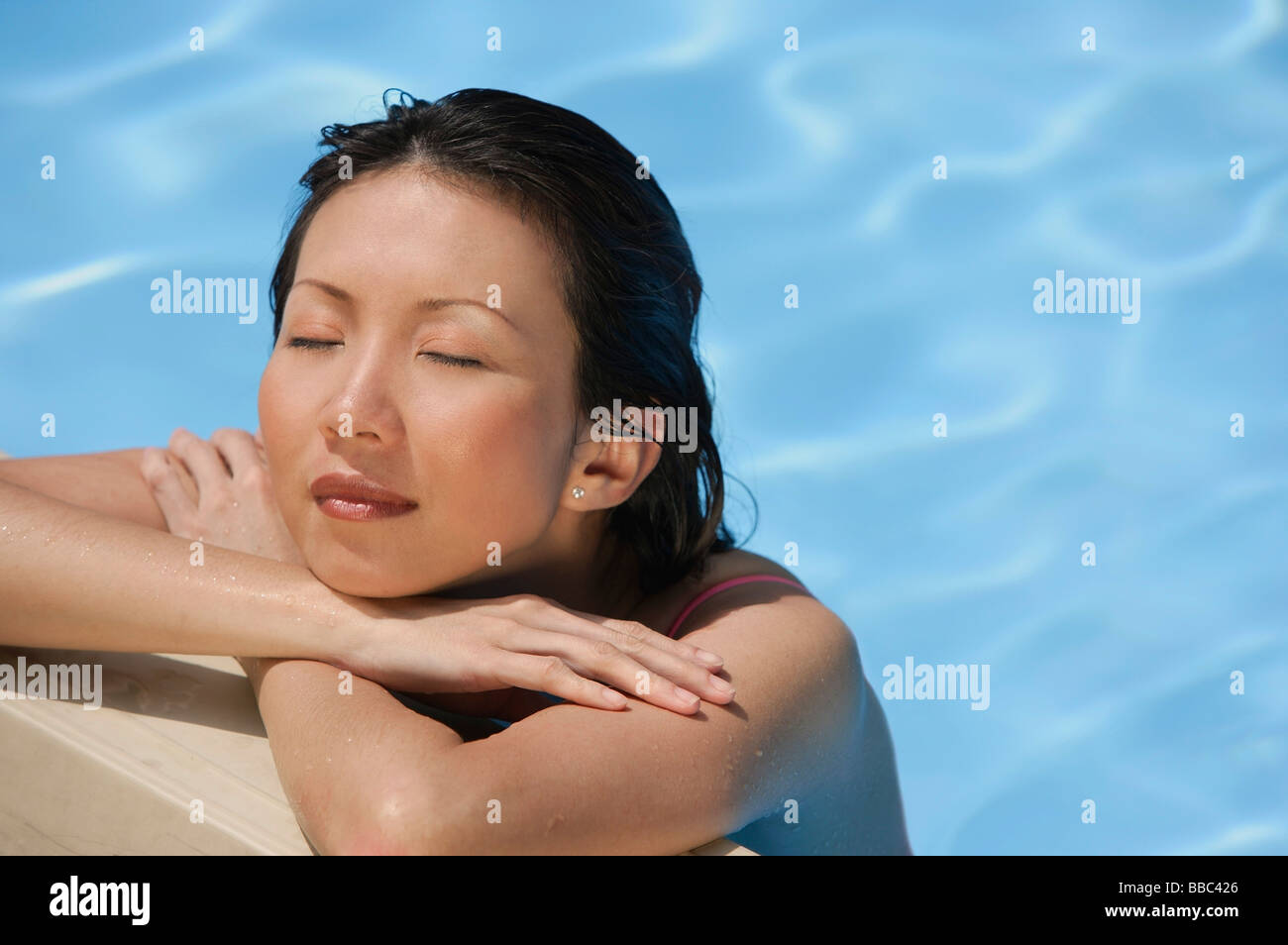 Woman leaning at the edge of swimming pool, arms crossed, eyes closed ...