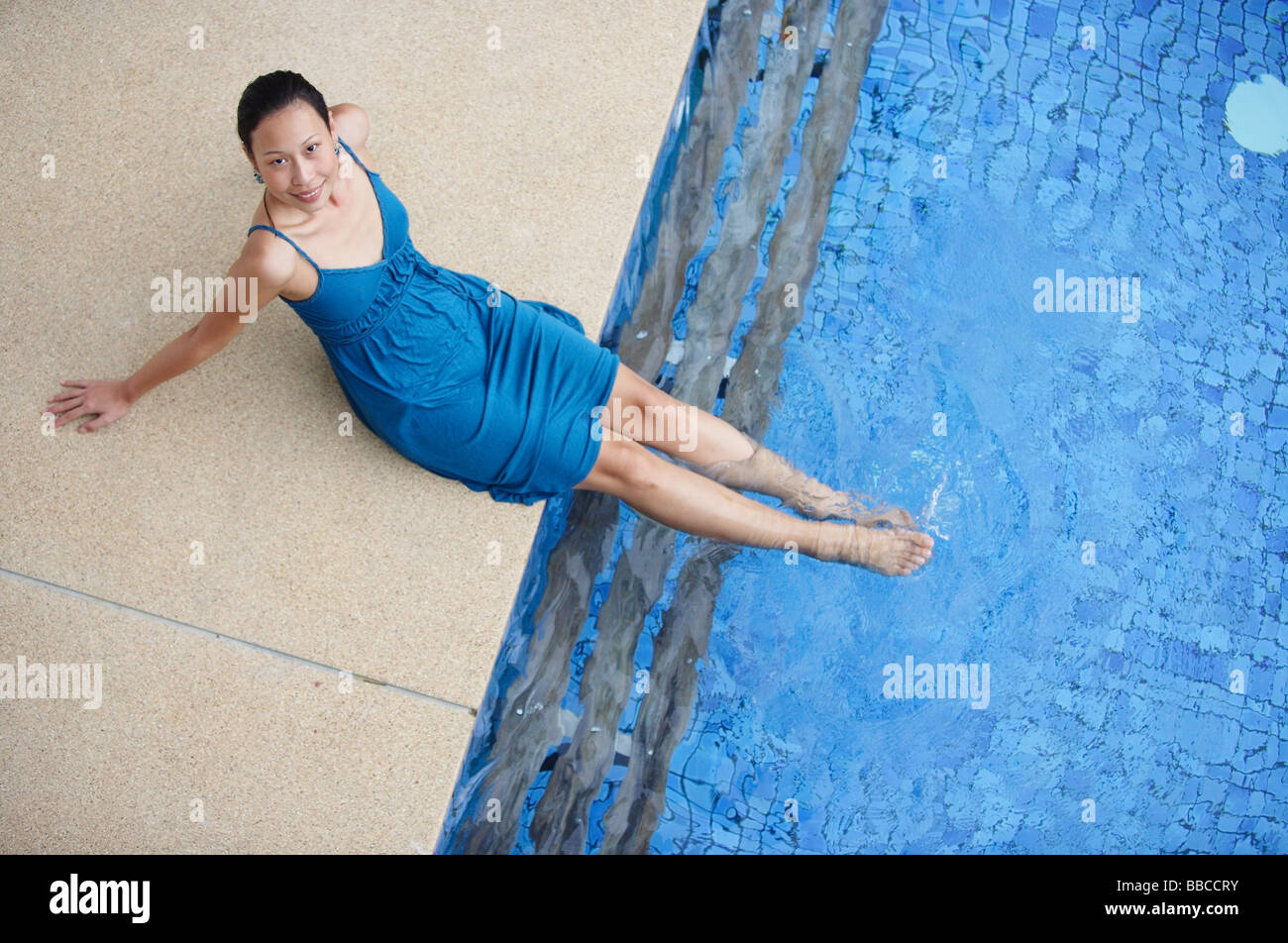 Young woman sitting by swimming pool, feet in water, smiling at camera ...