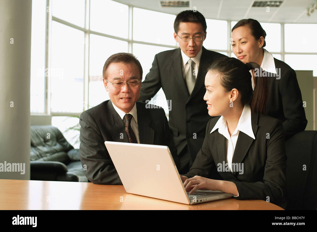 Executives in office, sitting and standing around open laptop, talking ...