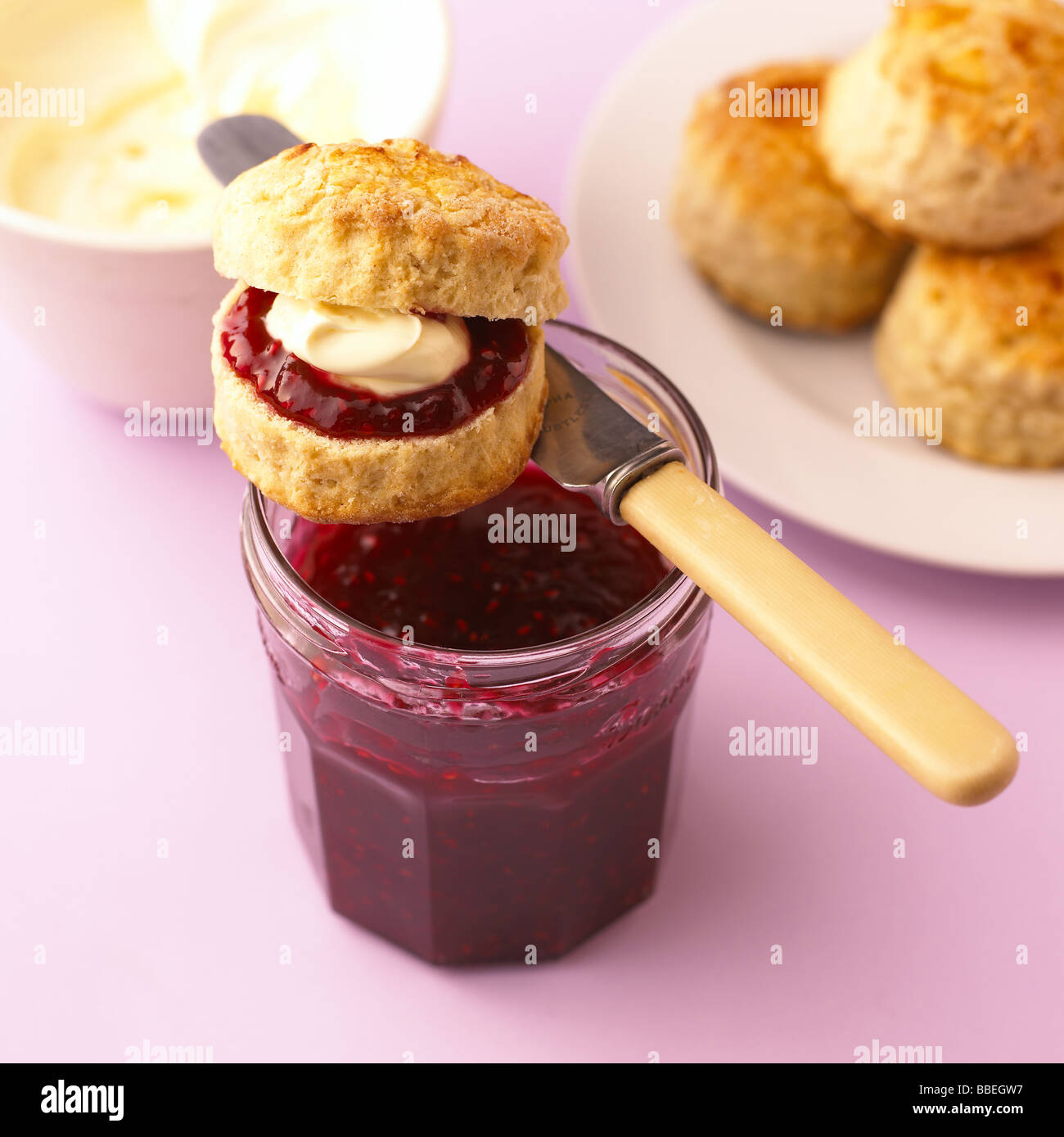 Scone filled with jam and cream resting on knife and jamjar on a pink ...