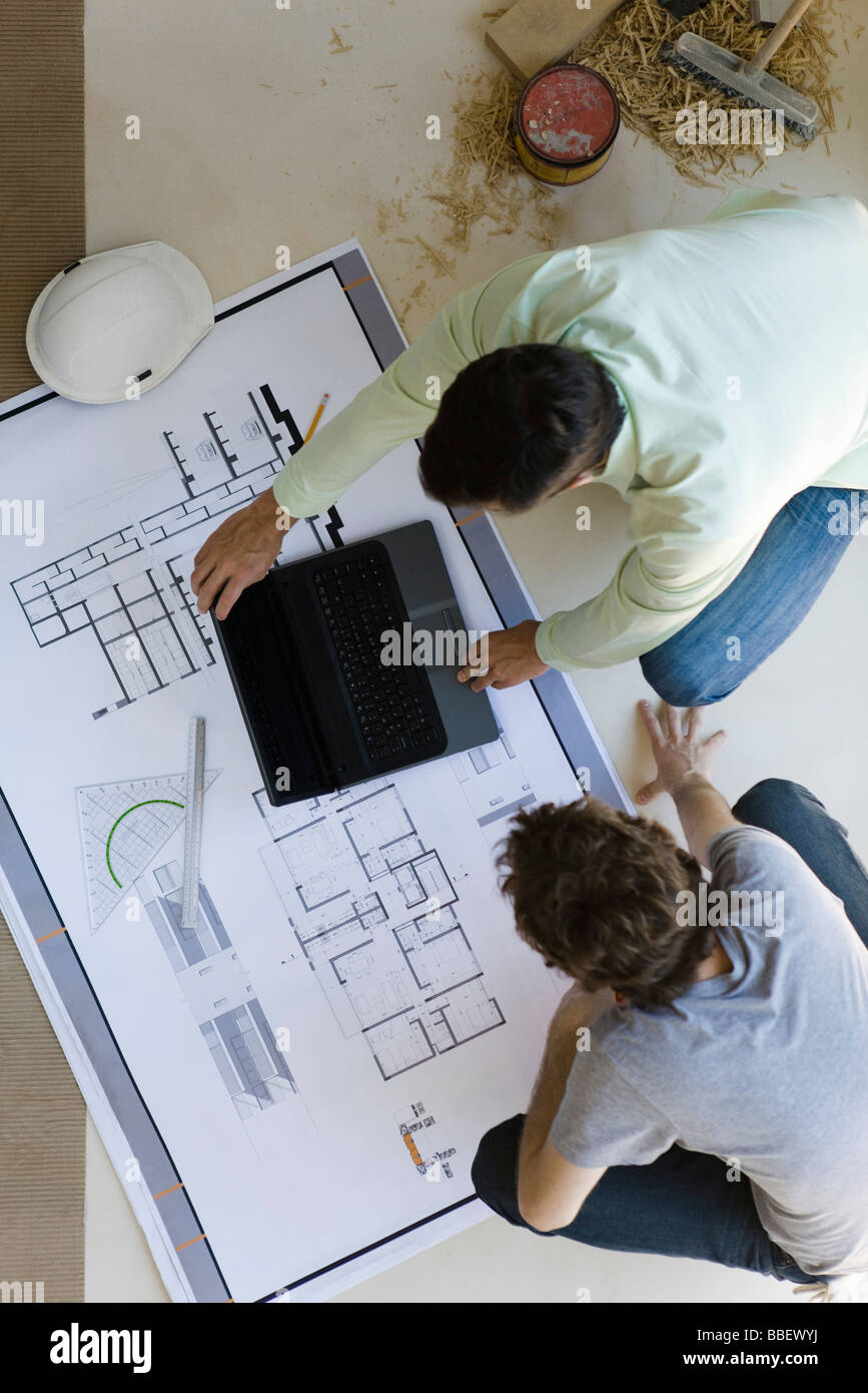 Two men crouching looking over blueprints, using laptop set on floor ...