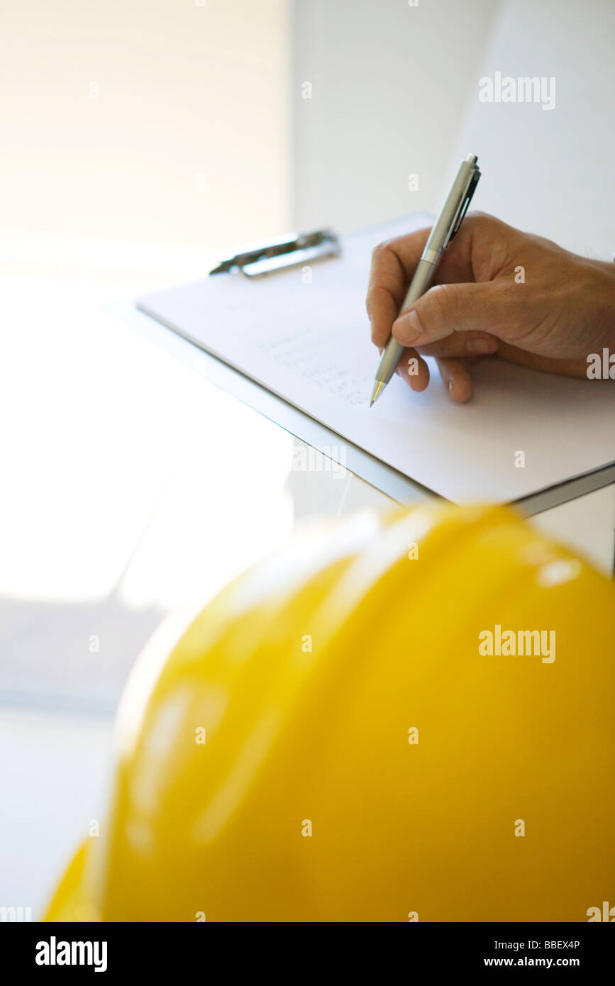 Hand writing notes on clipboard pad, hard hat in foreground, cropped ...