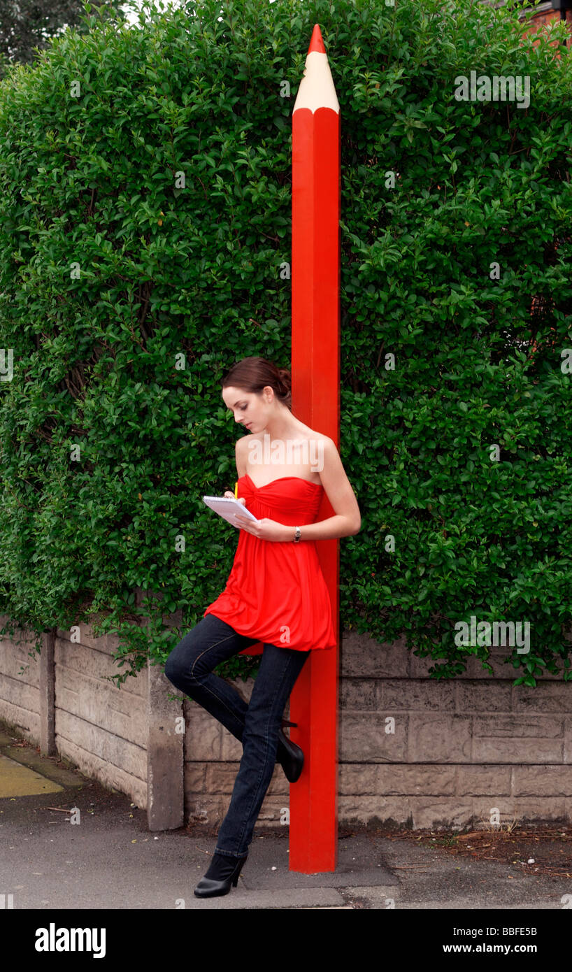 Young woman standing by giant-sized red pencil, writing on a notepad ...