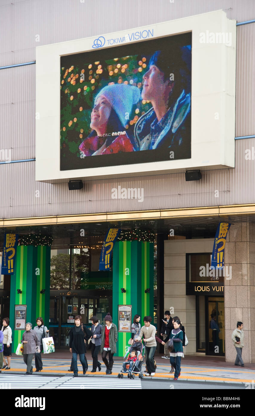 Pedestrians wait at crosswalk beneath large LCD screen above entrance ...