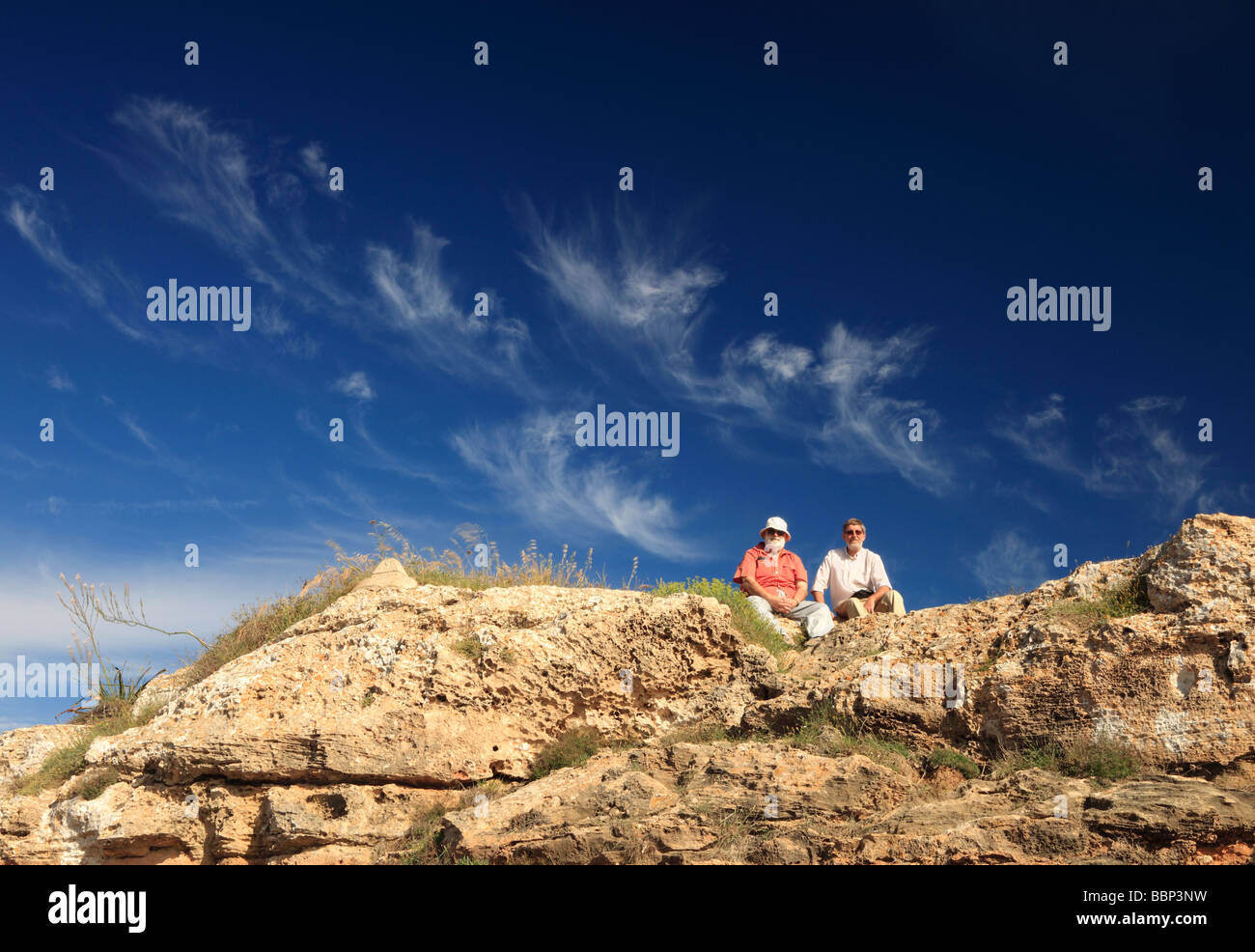Two men sitting on top of a rocky cliff with whispy clouds and a deep ...