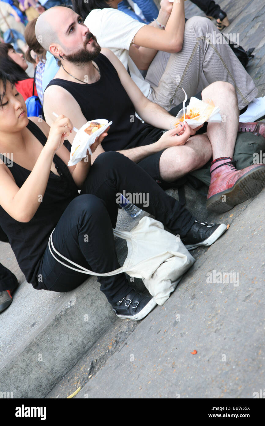 A couple having food, sitting on a pavement. Brick Lane, Sunday Market ...