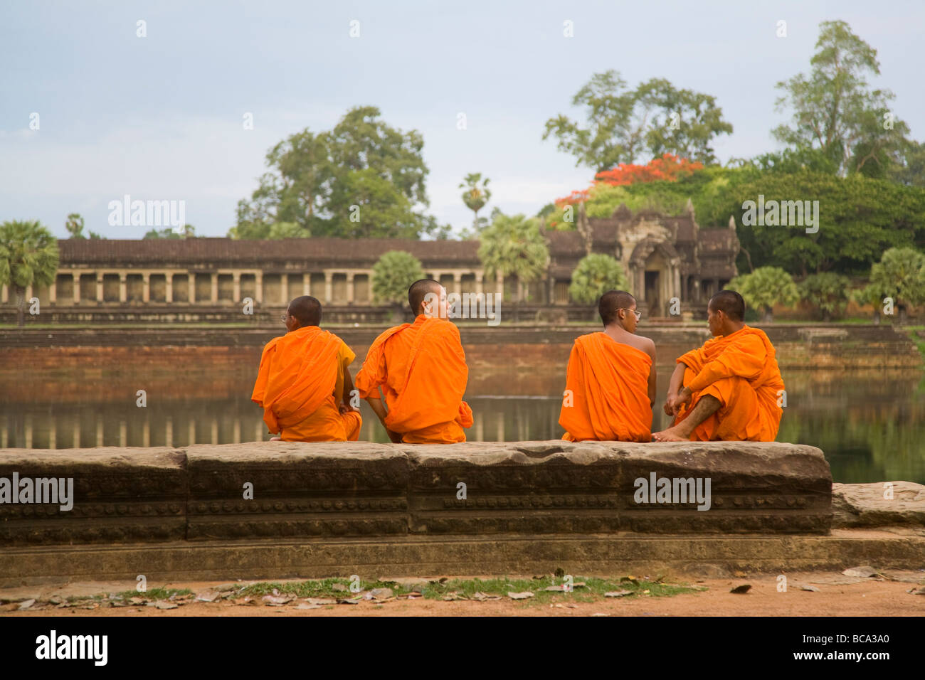 Four Khmer buddhist monks resting on stone wall of outer moat in Angkor ...