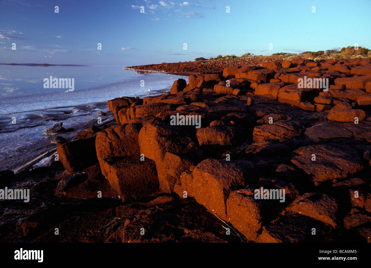 Volcanic red rocks line the shore of a flooded salt lake at sunset ...