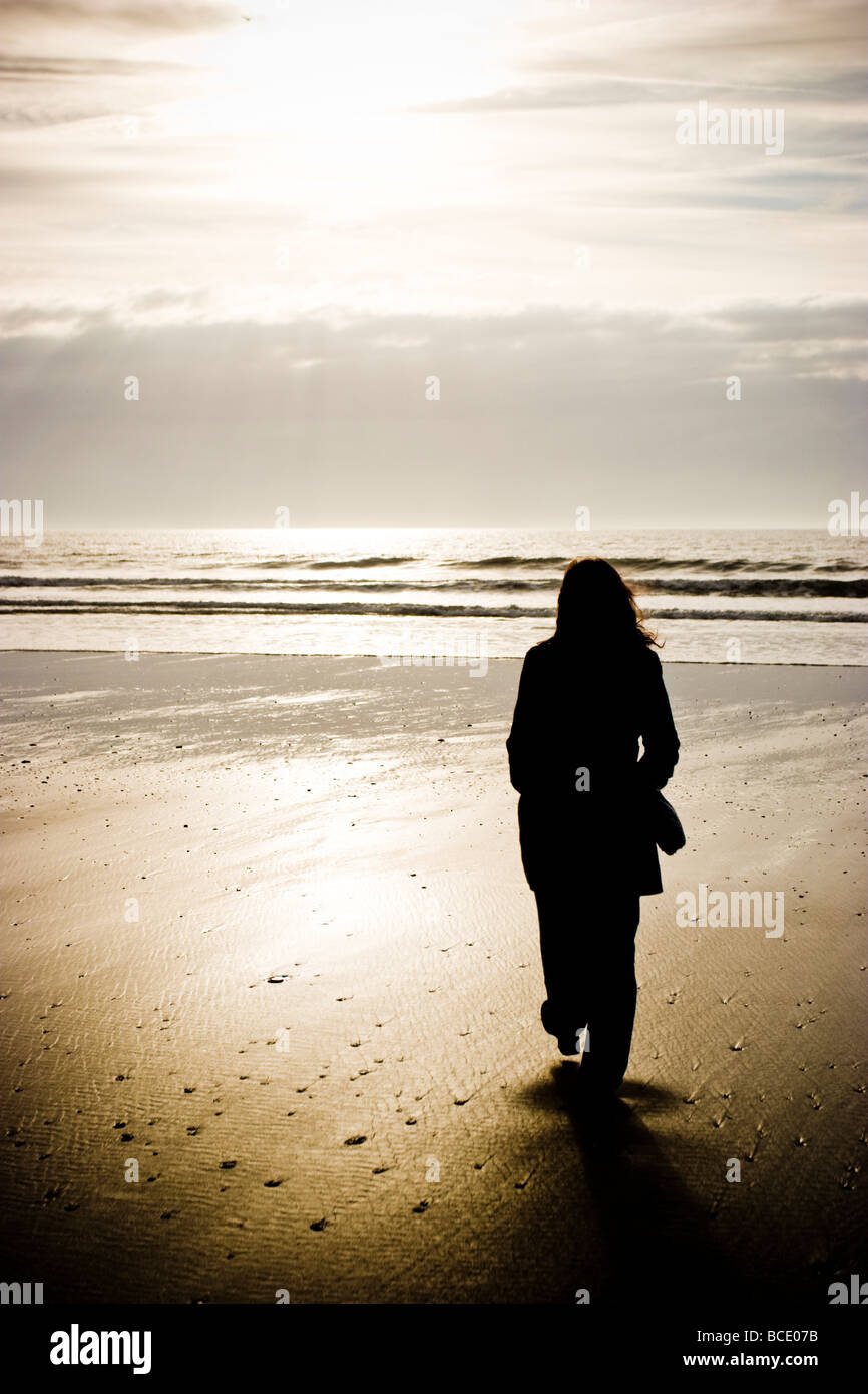 Woman walking alone along shoreline silohuetted against the low sun ...