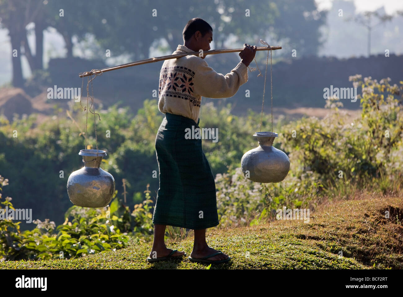 Myanmar, Burma, Mrauk U. A Rakhine man carries water in aluminium pots ...