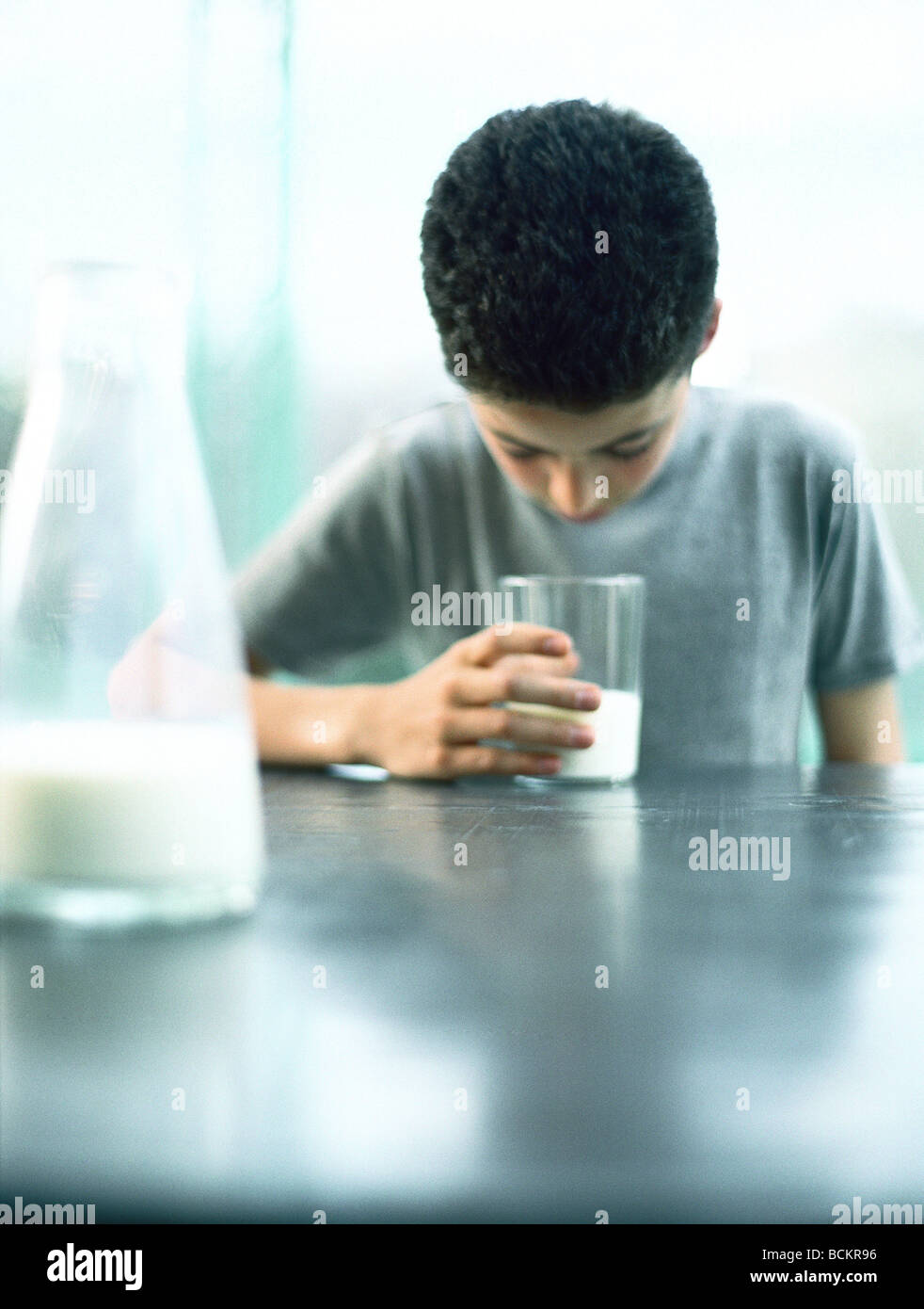 Boy sitting at table looking down into glass of milk, carafe of milk in ...