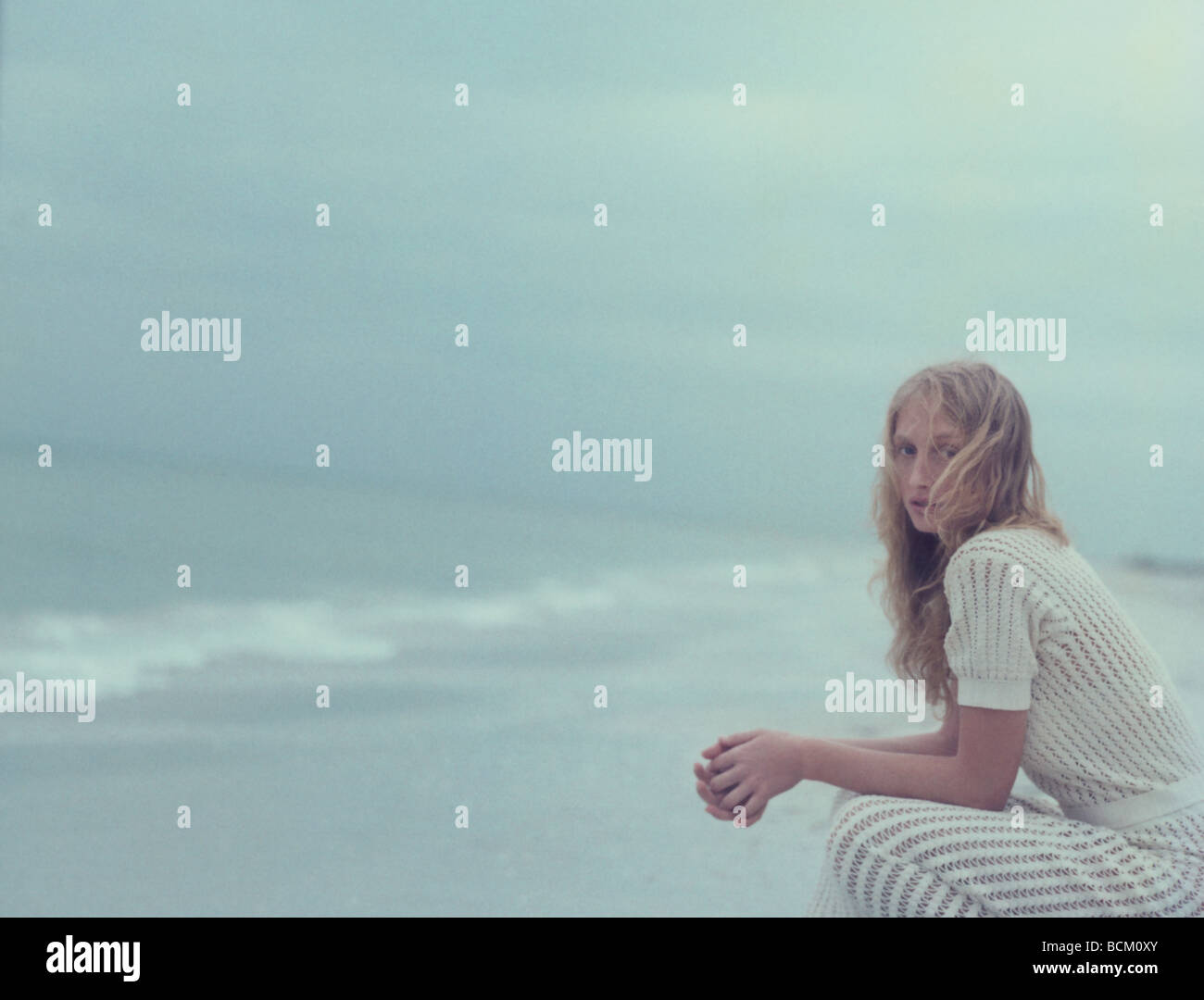 Young female in dress crouching on beach, looking at camera, side view ...