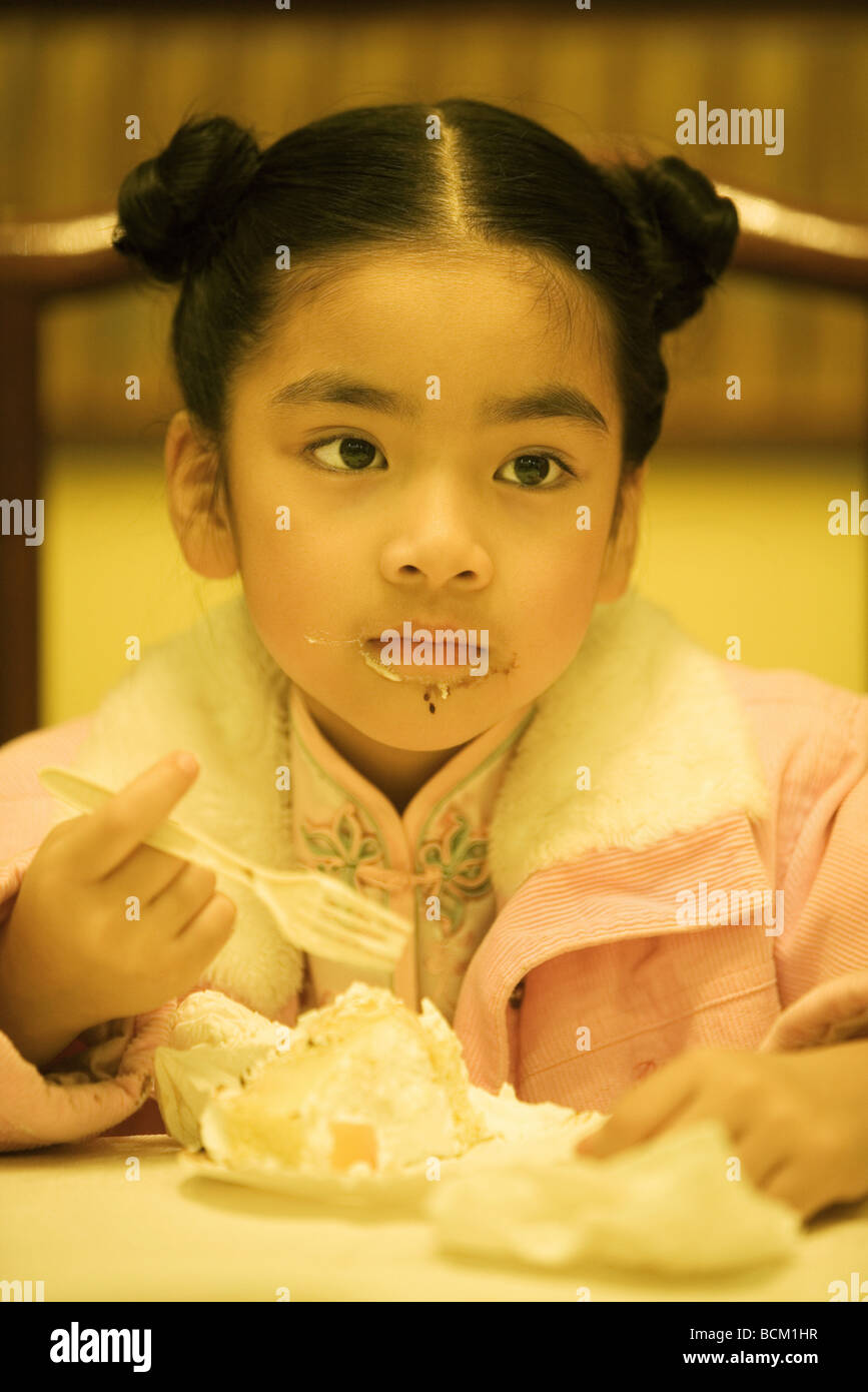 Girl sitting with fork poised over piece of cake, icing on face ...