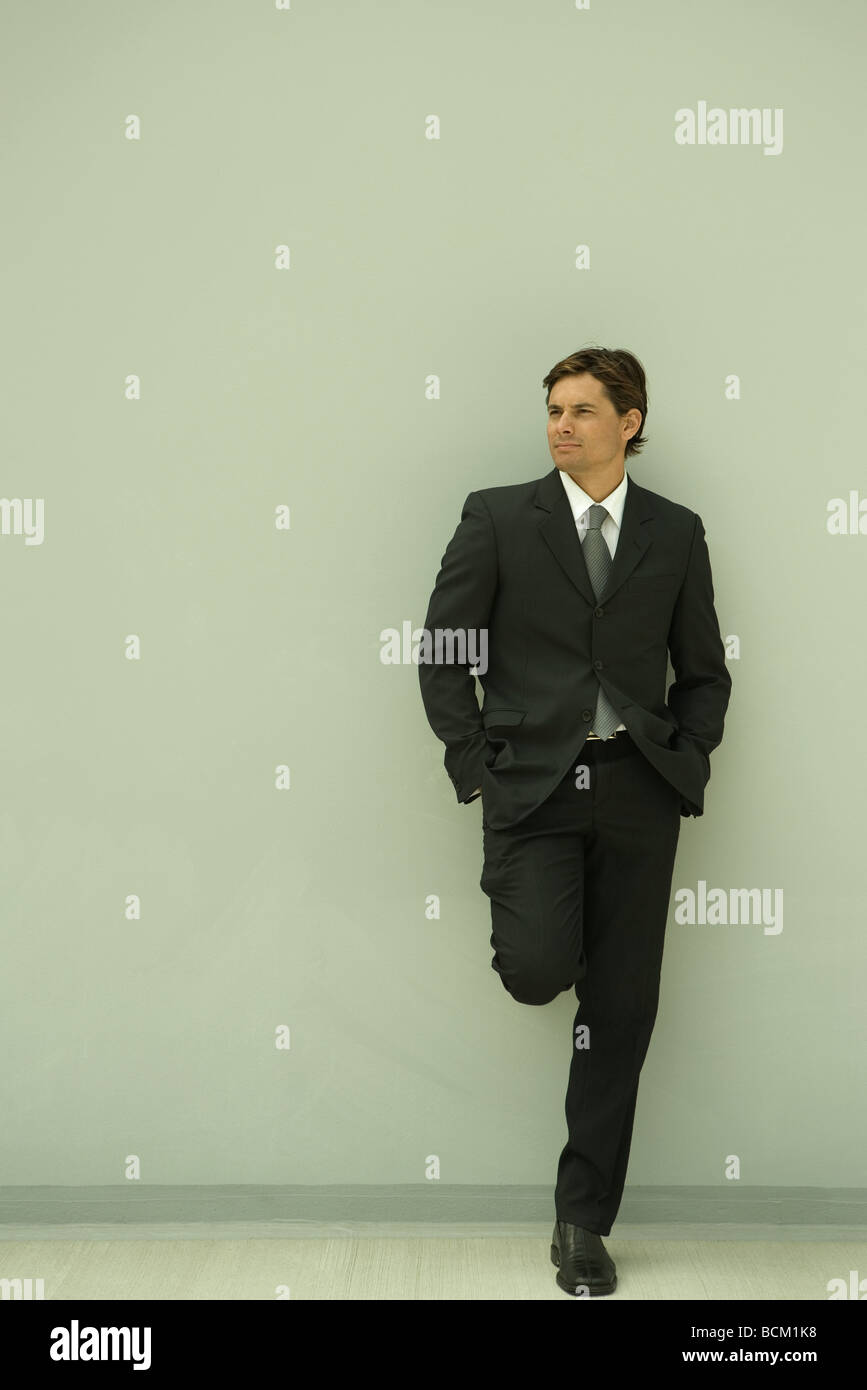 Businessman standing on one leg, leaning against wall with hands in ...