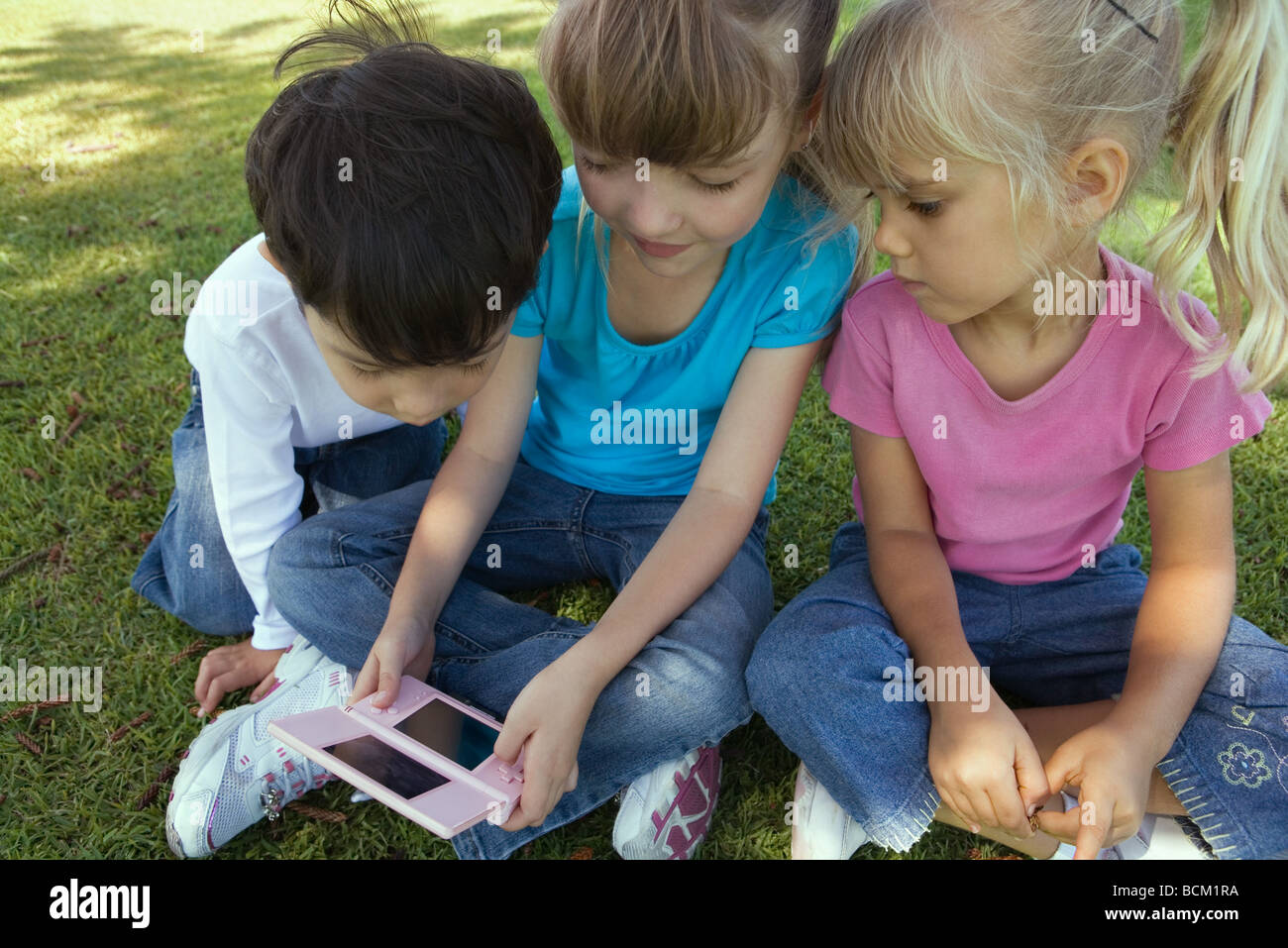 Three children sitting on grass, playing with video game, close-up ...