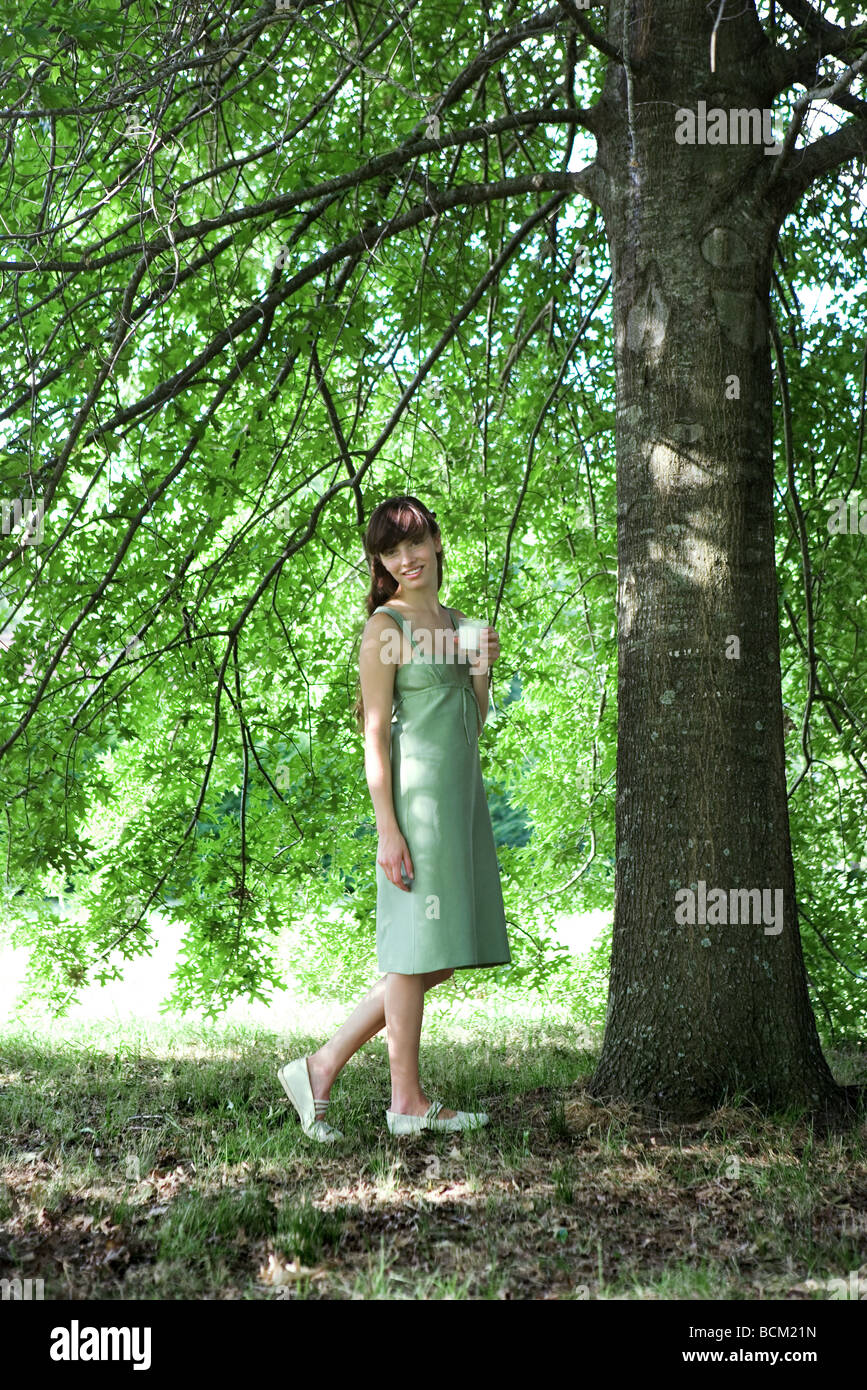 Young woman standing under tree, holding glass of milk, smiling at ...
