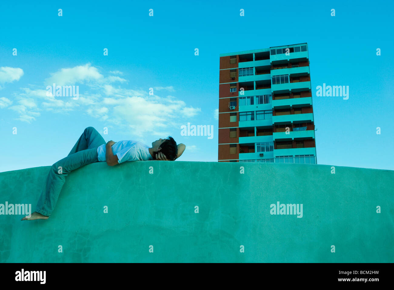 Young man lying on ledge, looking at sky, high rise apartment building ...