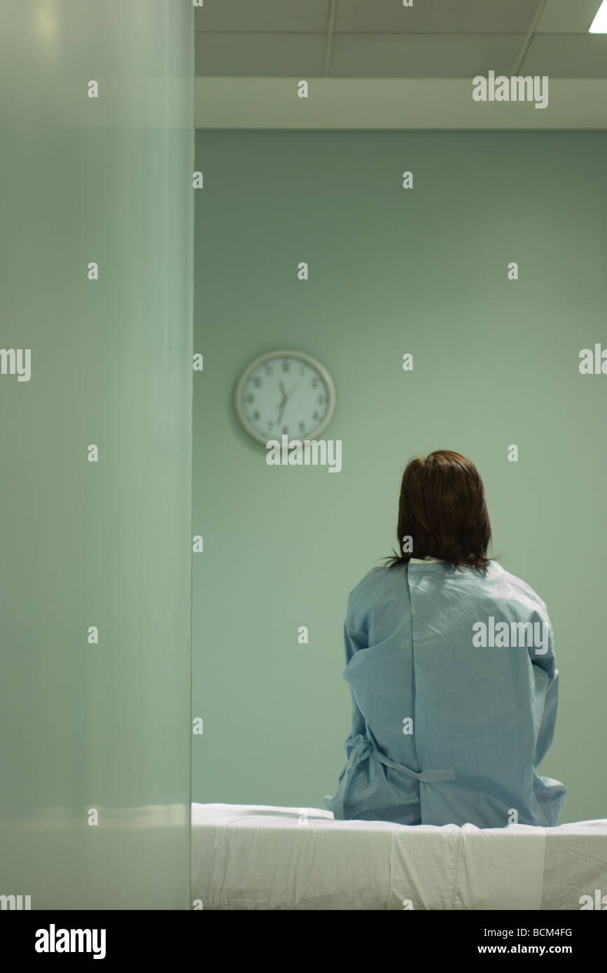 Female patient sitting on hospital bed, looking at clock, rear view ...