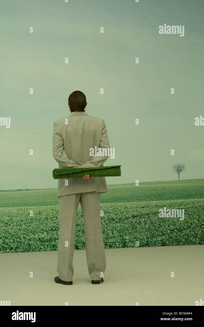 Man looking at rural scene, holding roll of artificial turf behind back ...