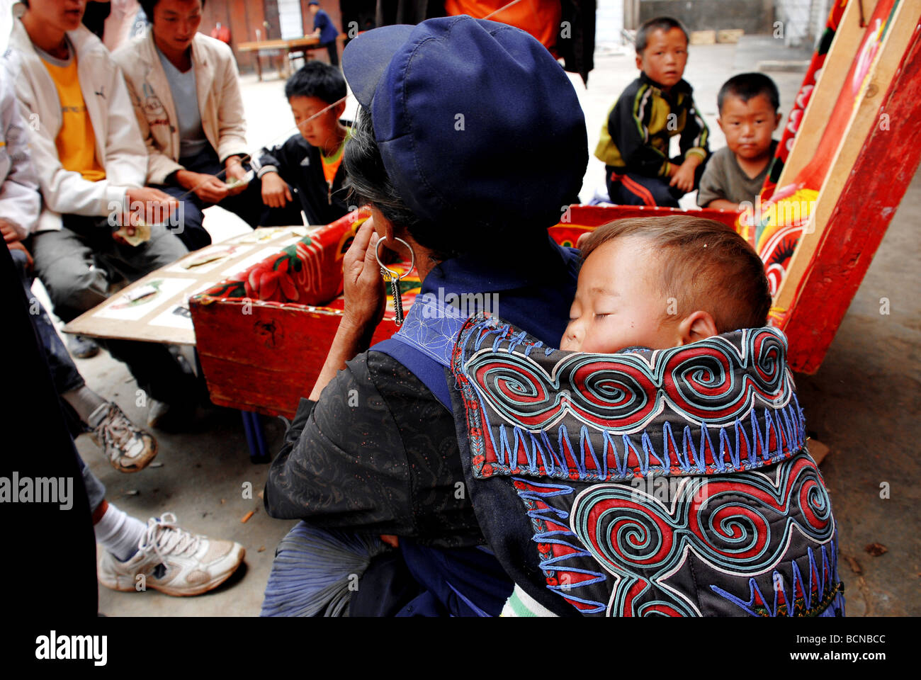 Yi minority woman carrying sleeping baby watching others gamble ...