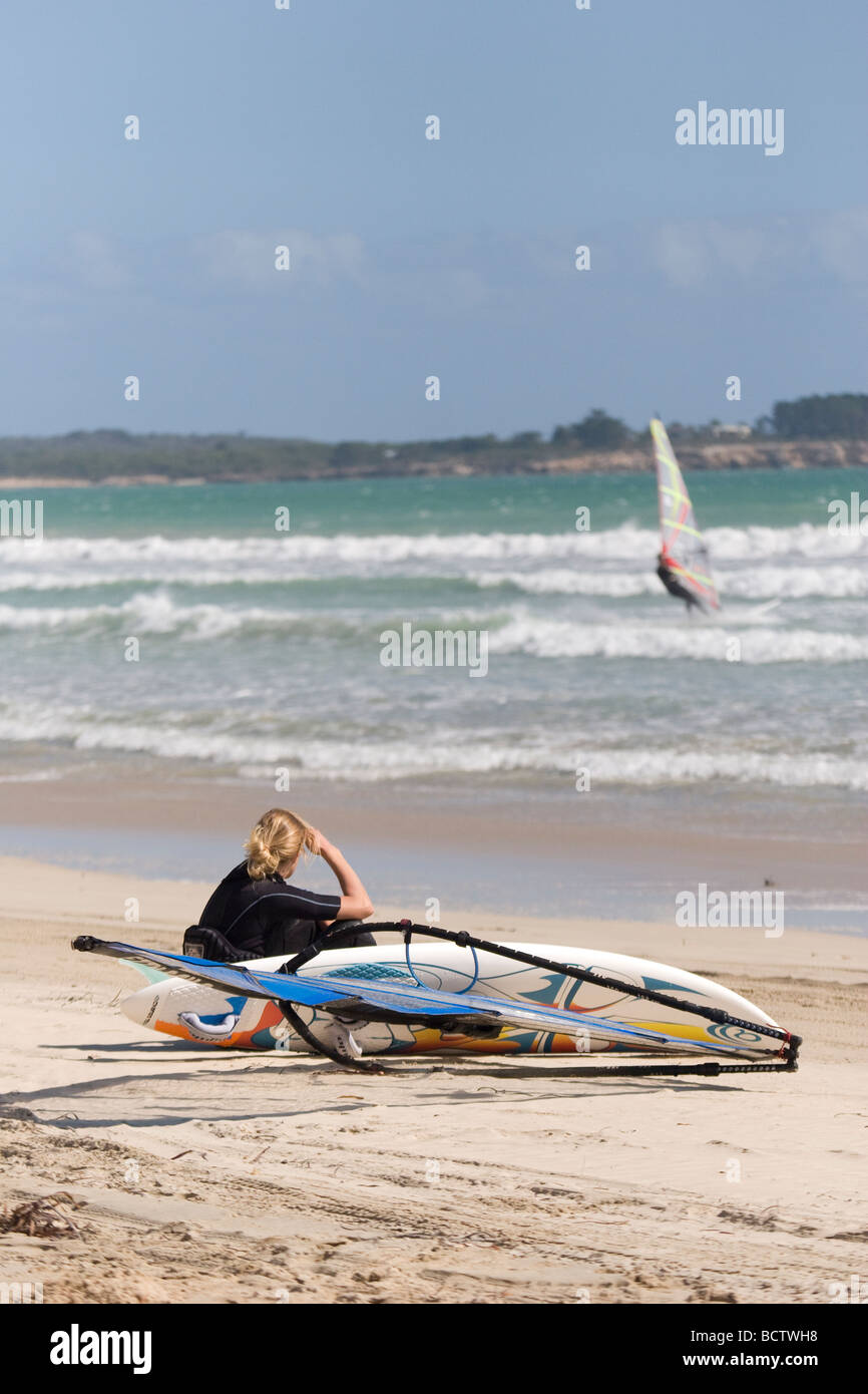 A windsurfer sits on the beach a watches another surfer out on the ...