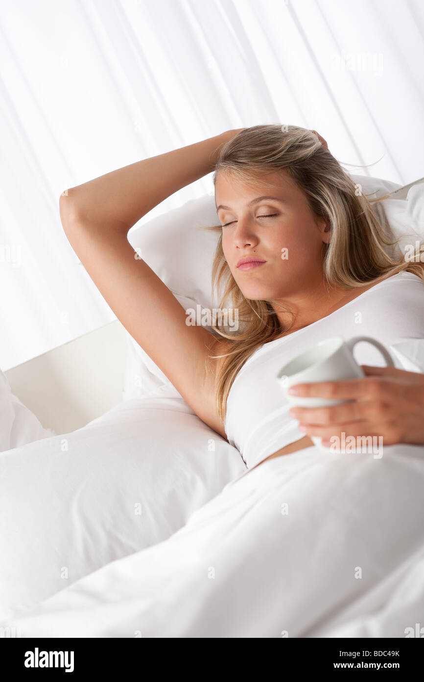 Blond woman holding white mug with coffee lying on white bed Stock ...