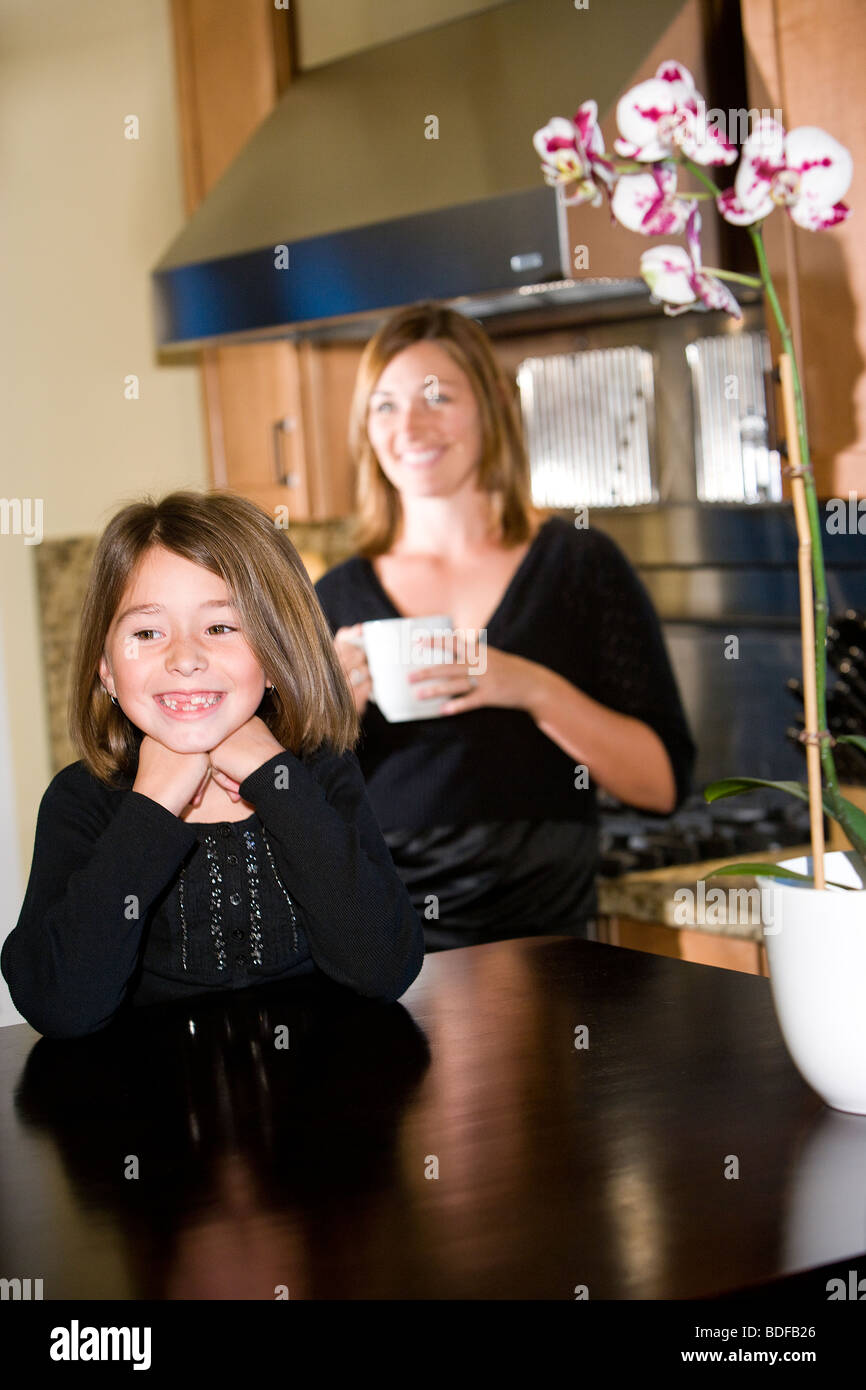 Little girl grinning with mother in background holding mug in kitchen ...