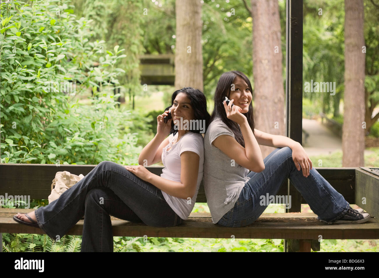 Two teenage girls sitting on a bench talking on their mobile phones ...