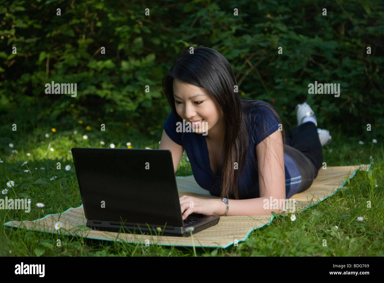 A young woman laying on a mat while using a wireless laptop in a park ...