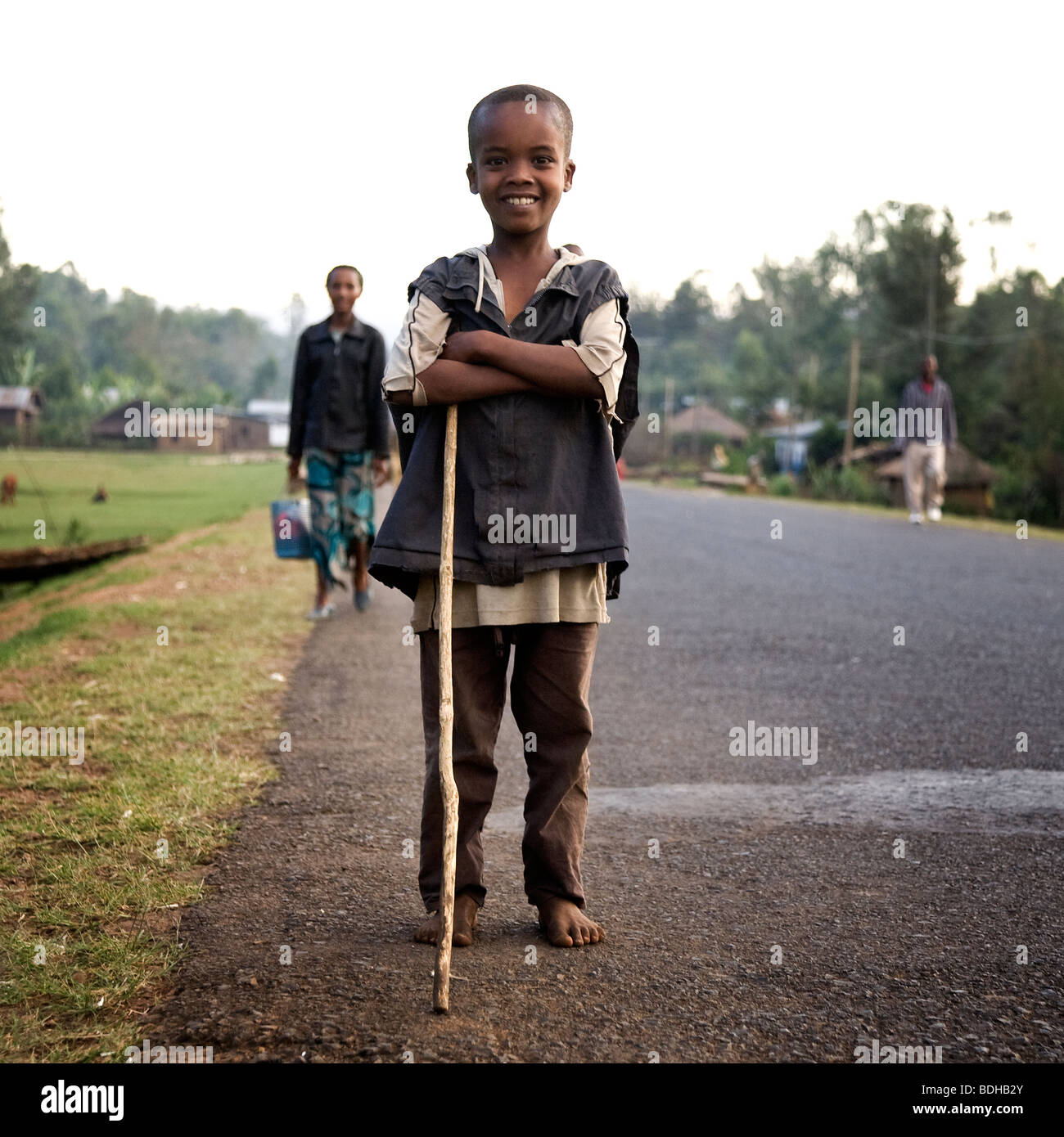 A boy with a walking stick poses on the edge of a paved road with ...