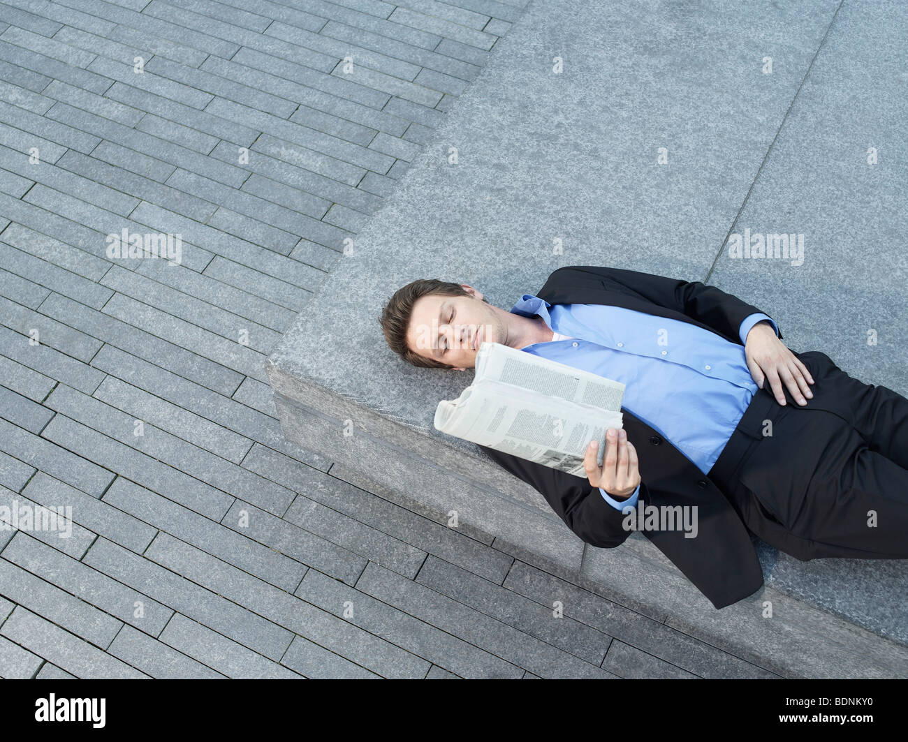 Young businessman lying on wall on pavement, reading newspaper ...