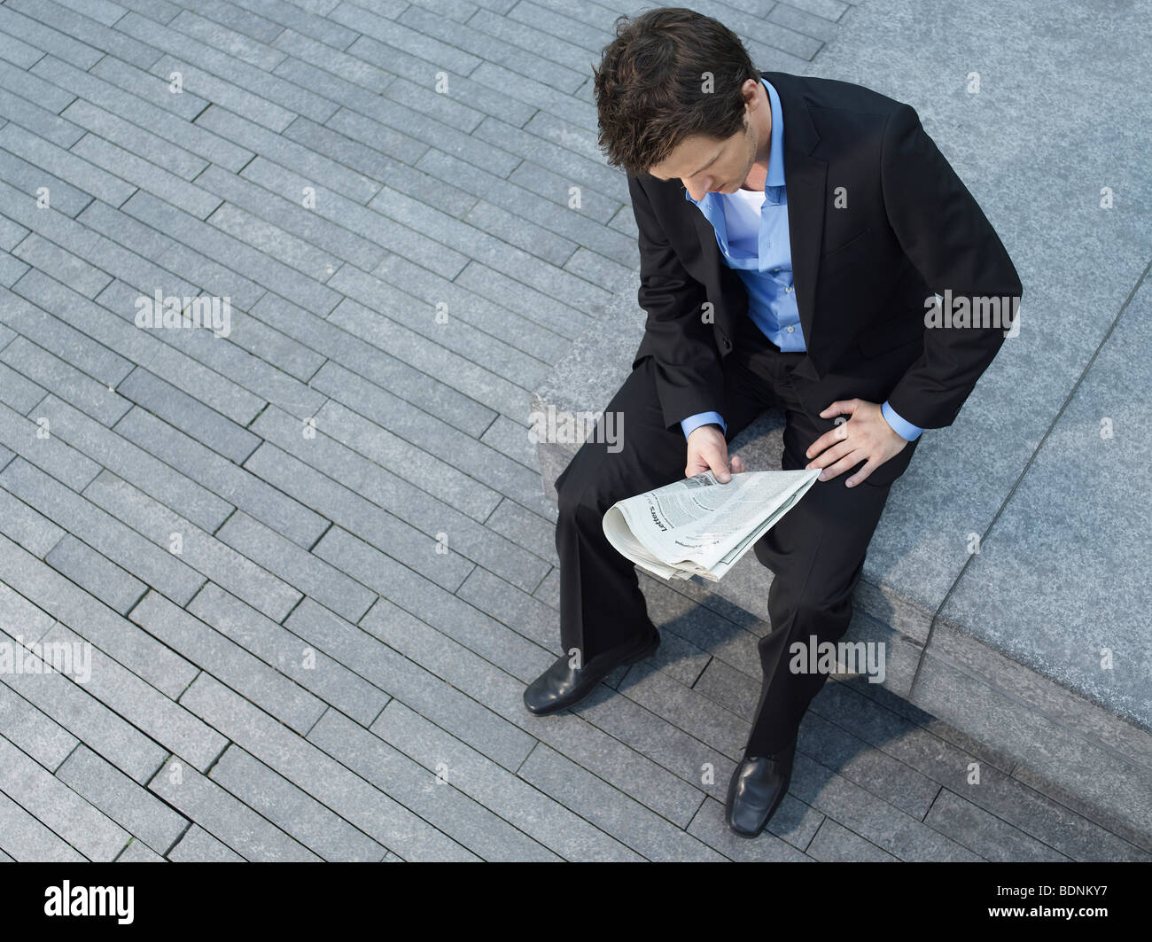 Young businessman sitting on wall on pavement, reading newspaper ...