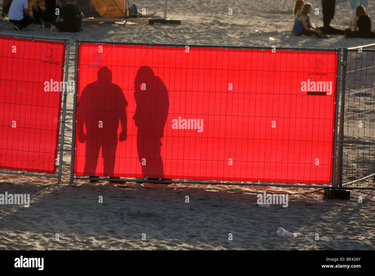Two men silhouettes behind red fence foil at rave music party beach ...