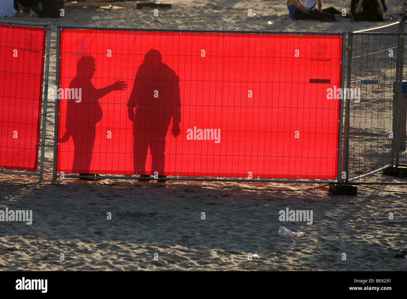 Two men silhouettes behind red fence foil at rave music party beach ...