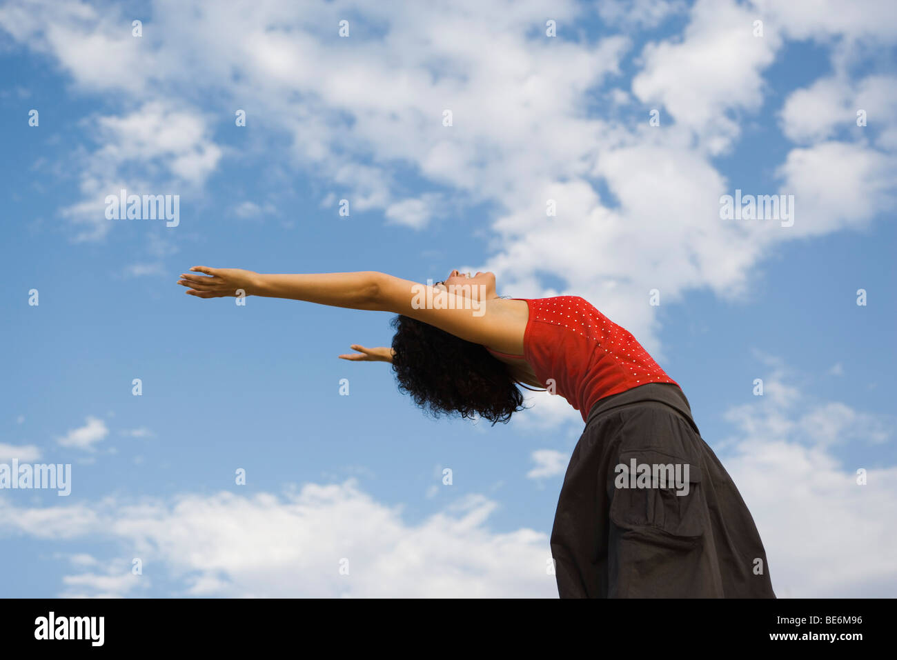 Young woman leaning backwards with arms extended, sky in background ...