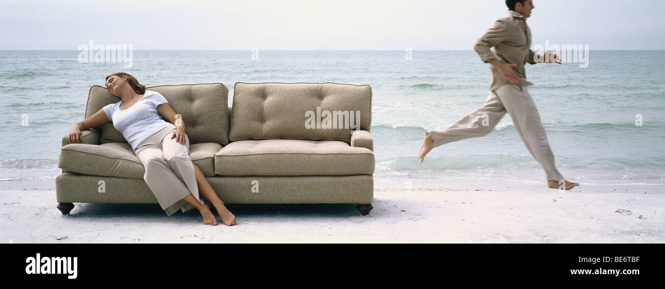 Woman napping on sofa on beach, man running past in background Stock ...