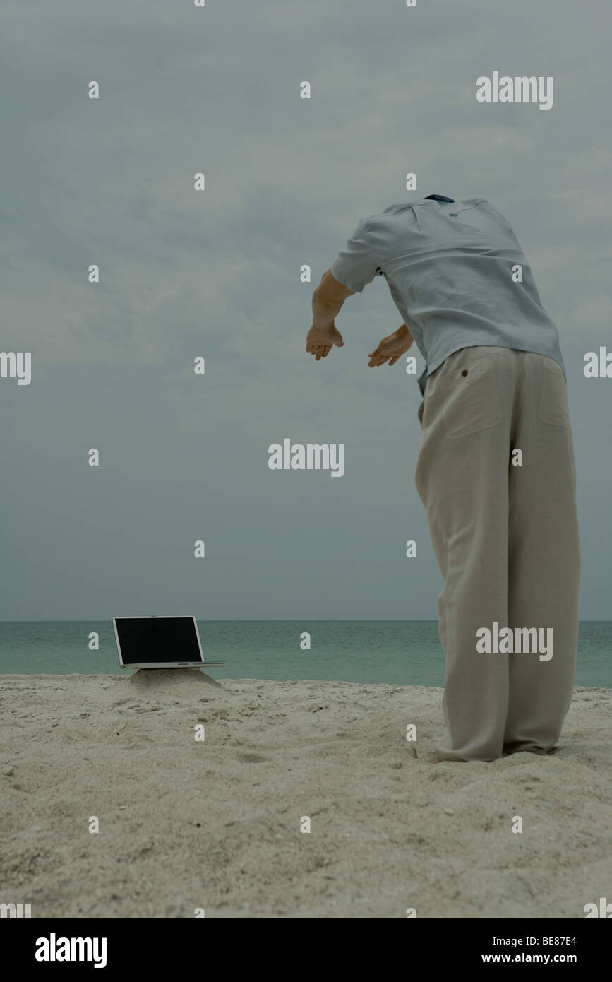 Man standing on beach, bending over toward laptop computer, rear view ...