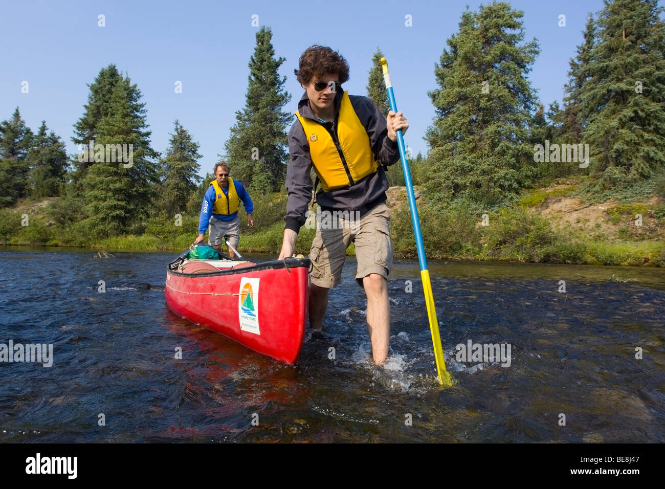 Two man lining canoe, walking bare feet in clear, shallow water of ...