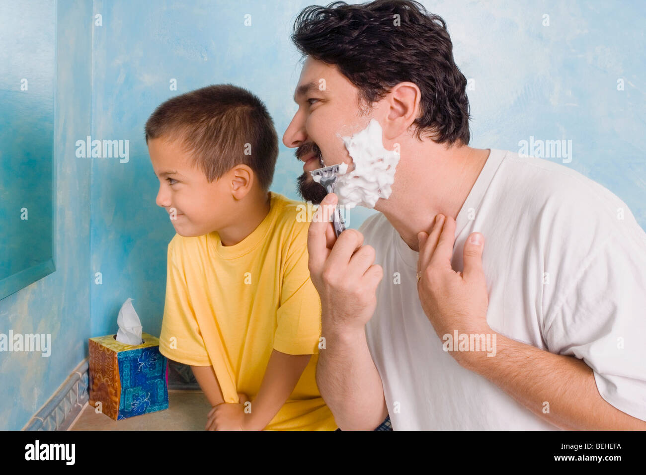 Side profile of a mid adult man shaving with his son sitting beside him ...
